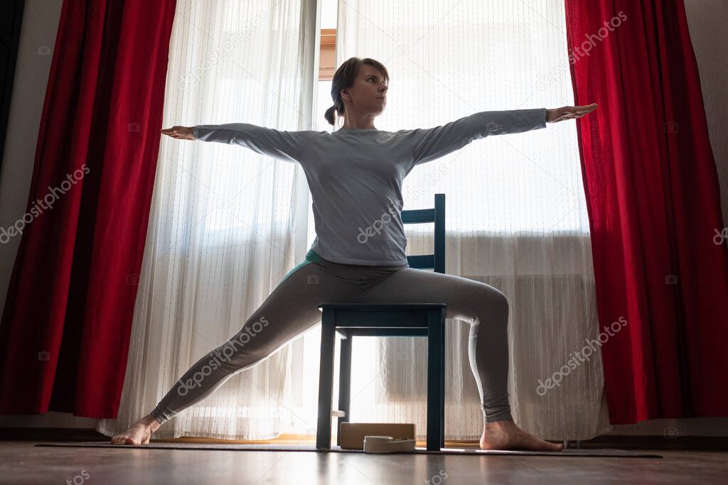 Mujer de yoga joven haciendo pose guerrera usando silla en la sala de ...