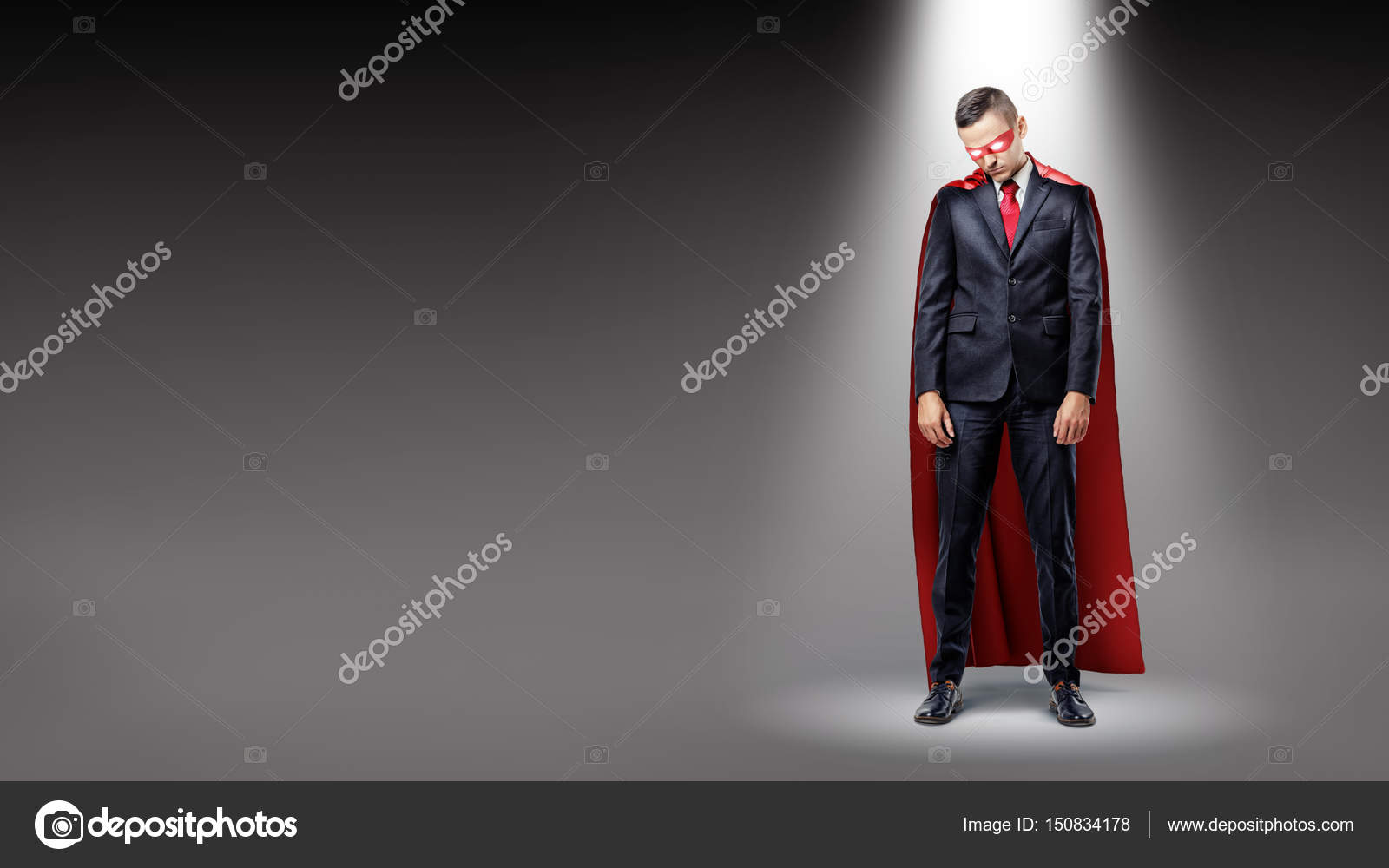 A sad businessman wearing a red superman cape standing in the spotlight ...