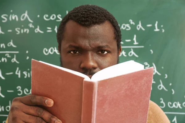 African-American math teacher with book near blackboard in classroom ...