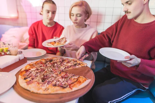 A group of friends sits in a pizzeria and eats a big pizza. Young ...