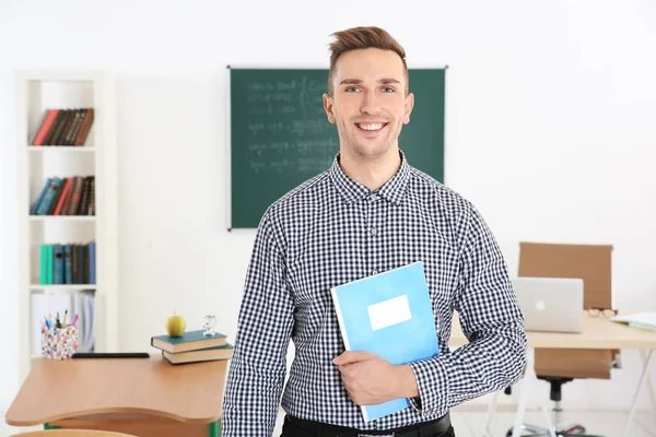 Young male teacher with book sitting at table in classroom Stock Photo ...