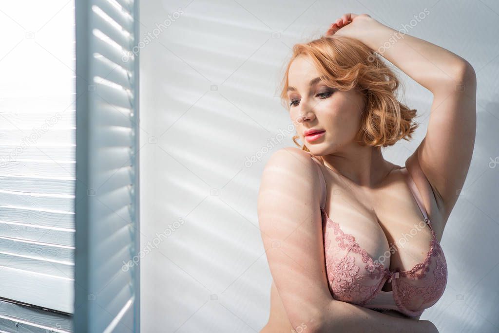Una hermosa joven con senos naturales muy grandes está de pie junto a la ventana con persianas ...