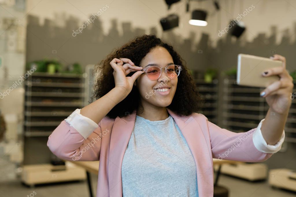 Chica morena sonriente con tel fono inteligente haciendo selfie en la tienda de ptica al probar ...