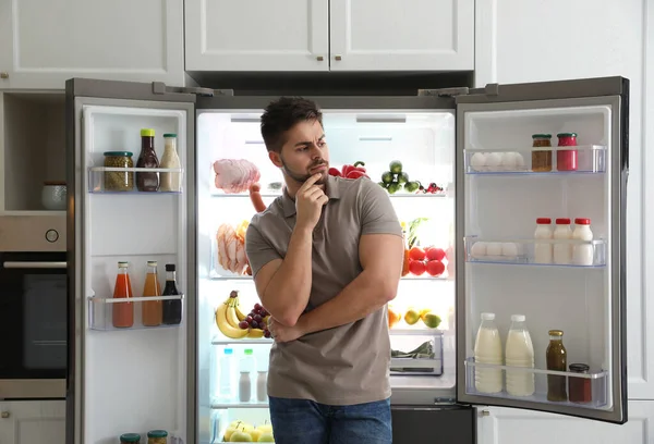 Thoughtful young man near open refrigerator in kitchen - Stock Image ...