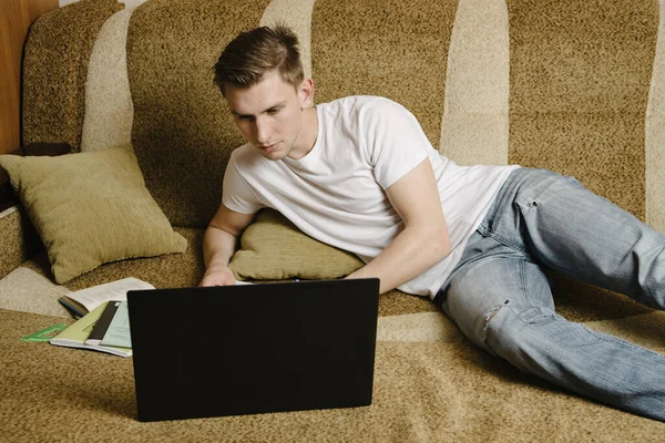 A young male student lying on a couch and using laptop while doing ...