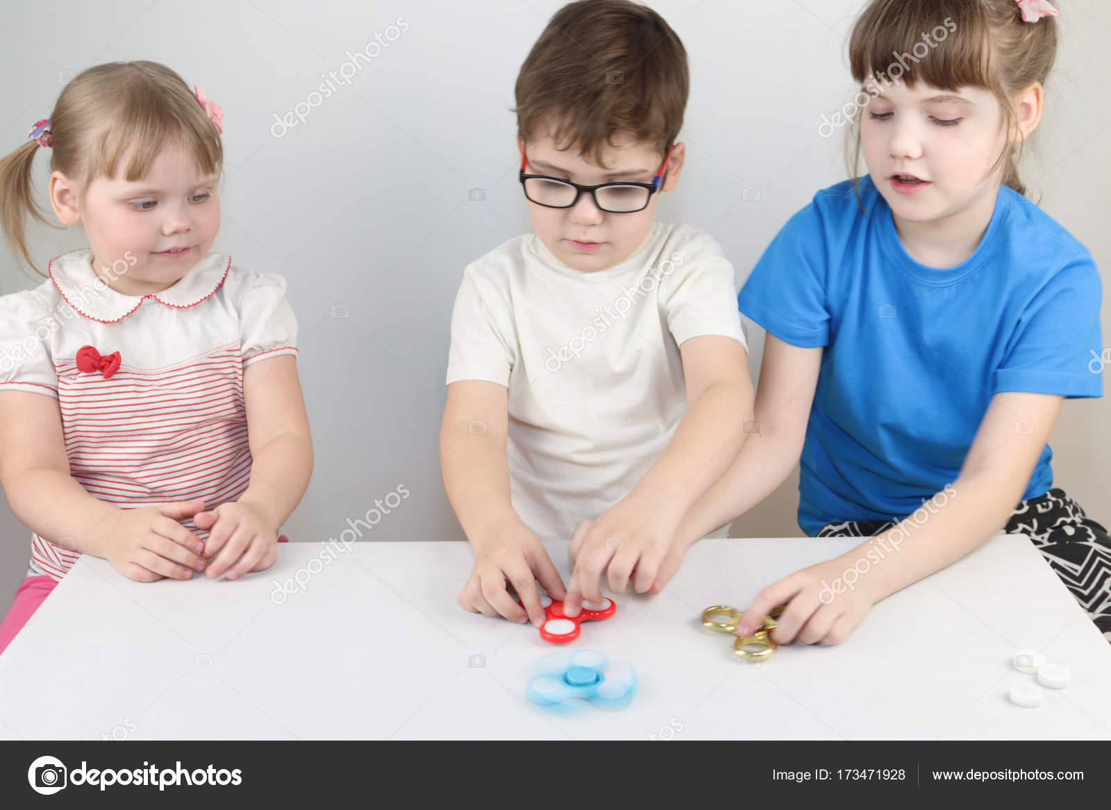 Two happy girls and boy play with spinners on table in white stu ...