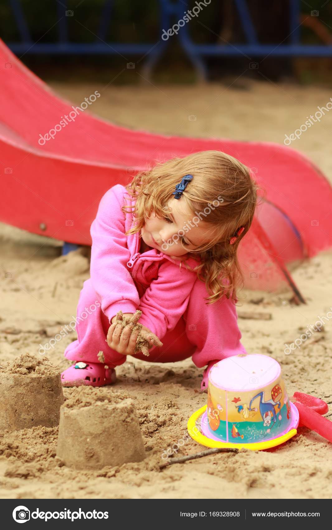 One year old cute curly girl in pink sports suit playing and making sand castles on the ...