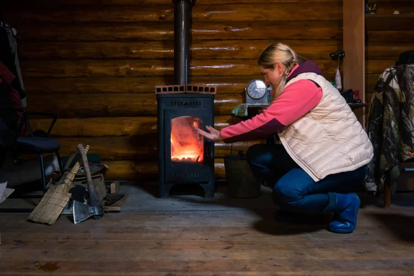 A woman crouched by a cast-iron stove in a wooden house and warms her ...