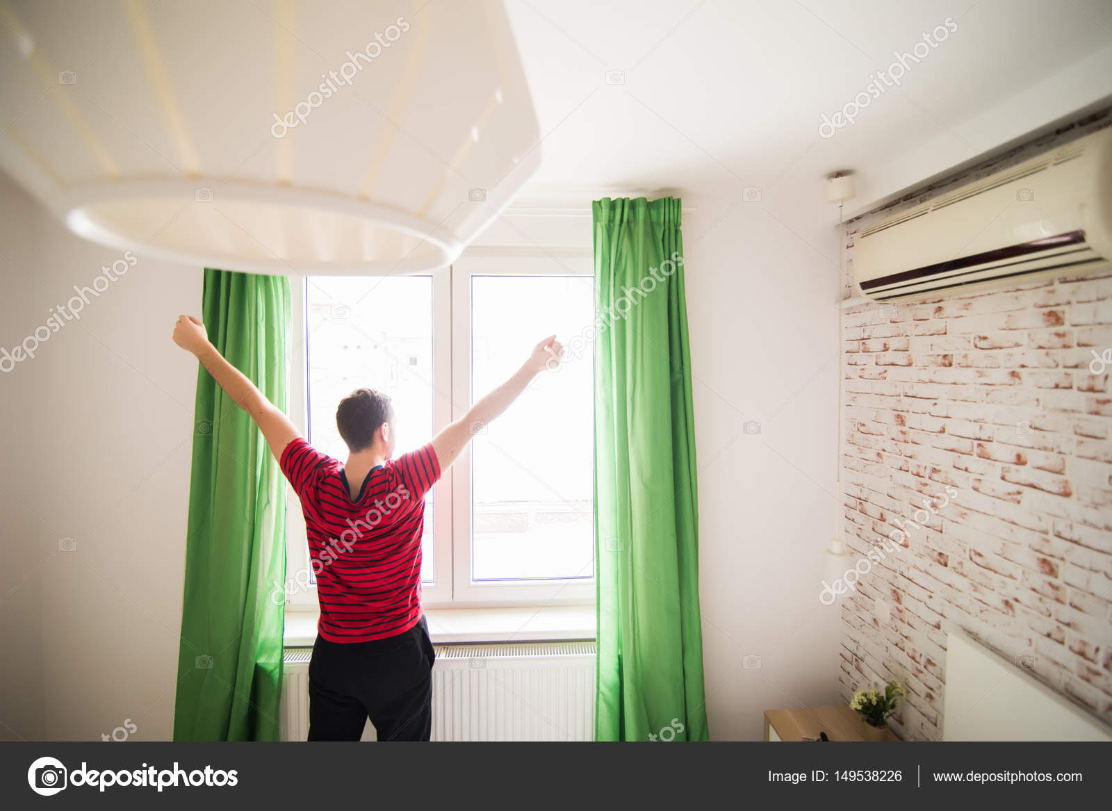 Handsome man waking up in the bed rising hands to window in the morning ...