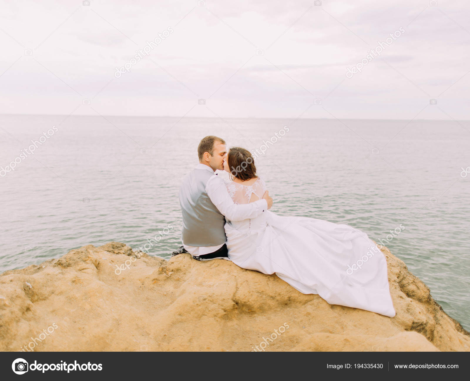 Romantic back portrait of the kissing couple while sitting on the cliff ...
