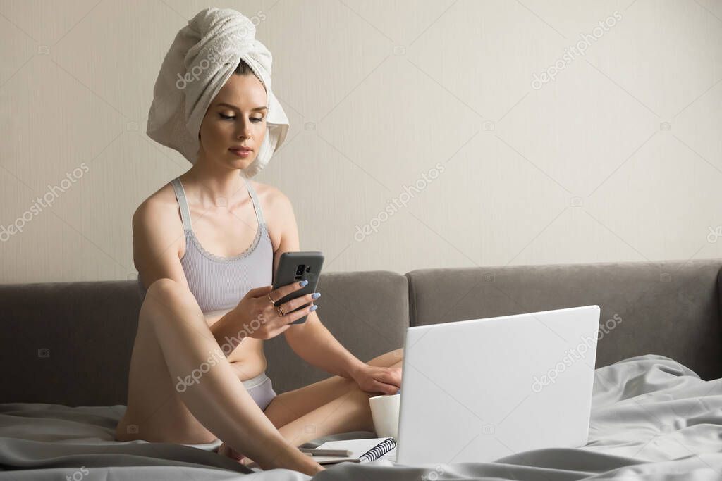 Mujer joven y bonita con una toalla en la cabeza se encuentra en la cama sostener el teléfono ...